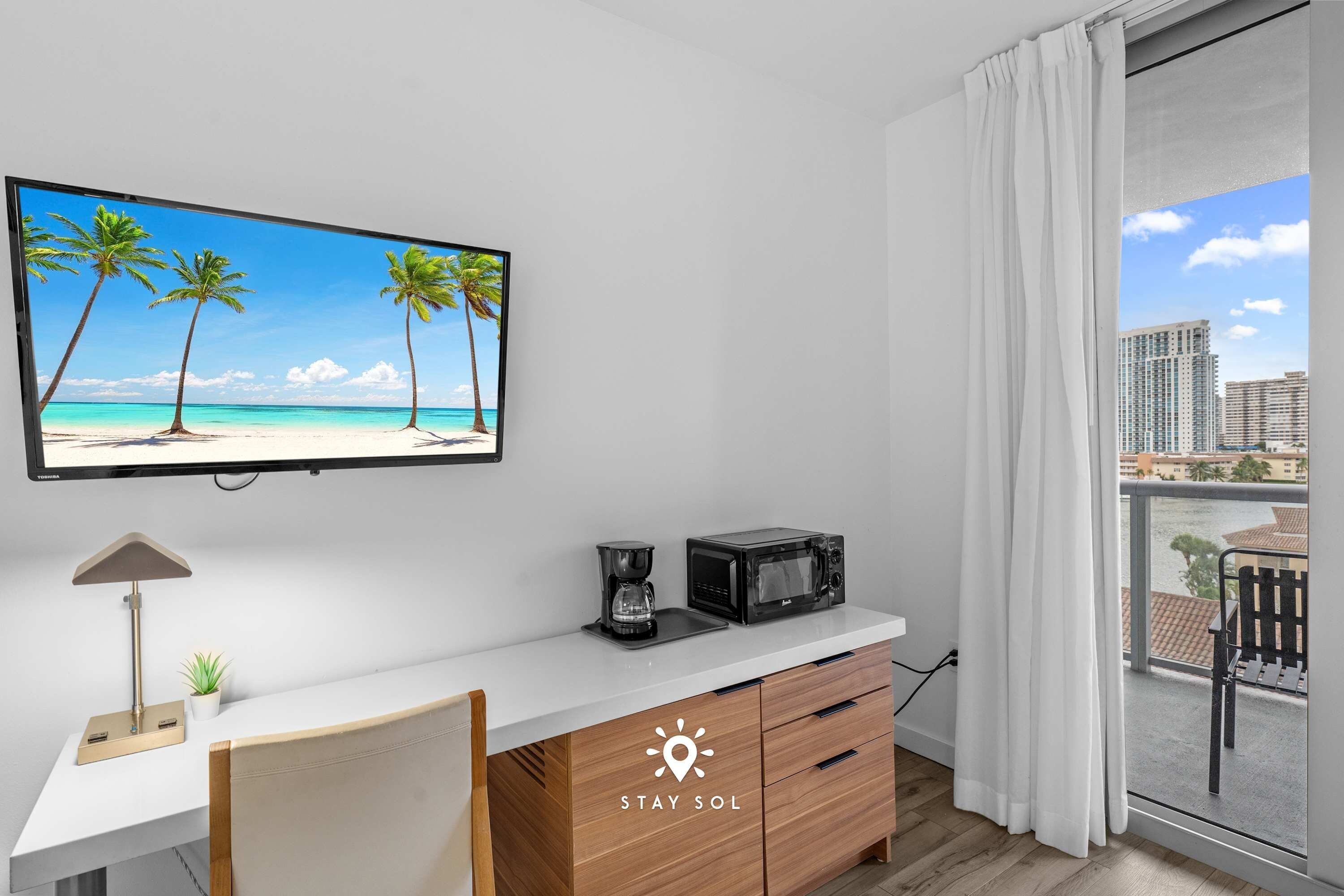 Quarto moderno com TV de tela plana exibindo uma cena de praia, cafeteira, micro-ondas e armário de madeira com o logotipo 'STAY SOL'.