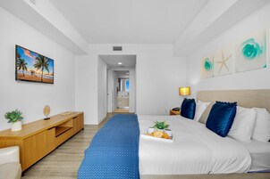Quarto de hotel com uma cama, mesa de cabeceira com abajur, televisão na parede e corredor que dá para um banheiro.