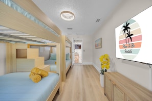 Quarto de hotel com beliches, corredor, banheiro e parede com pôster de tema tropical.