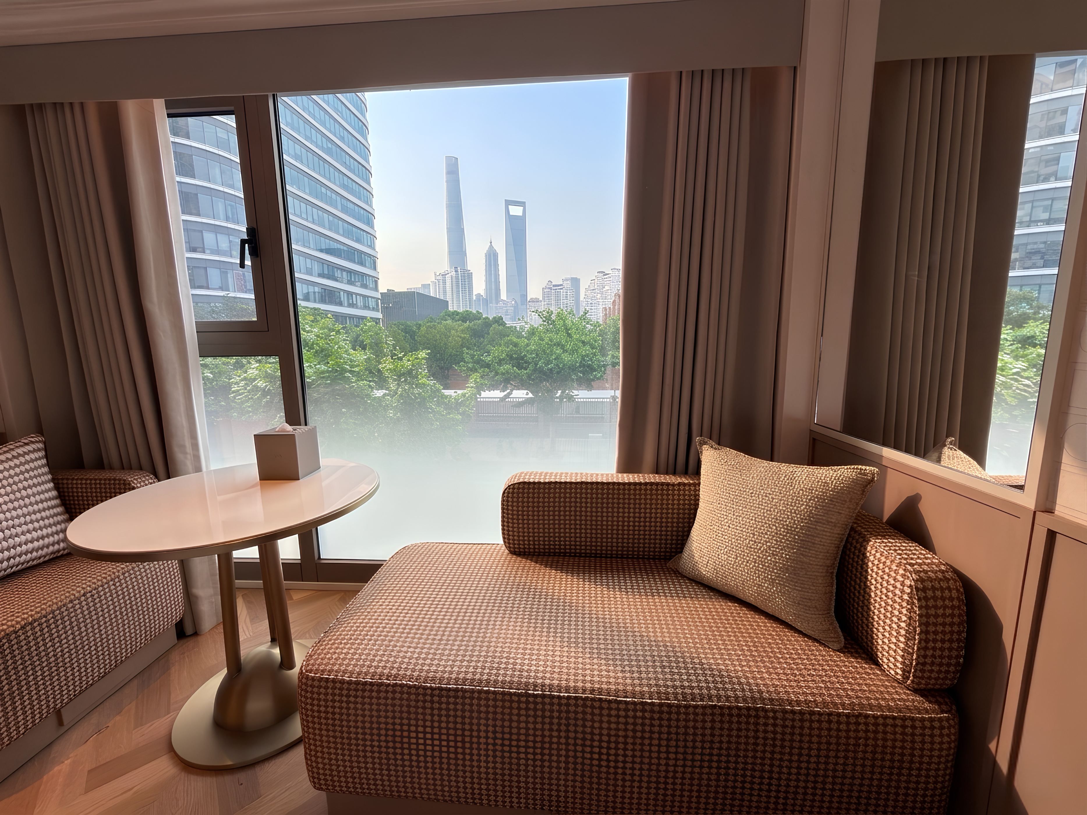 Quarto de hotel moderno com uma grande janela, um sofá, uma mesinha e vista para a paisagem urbana.