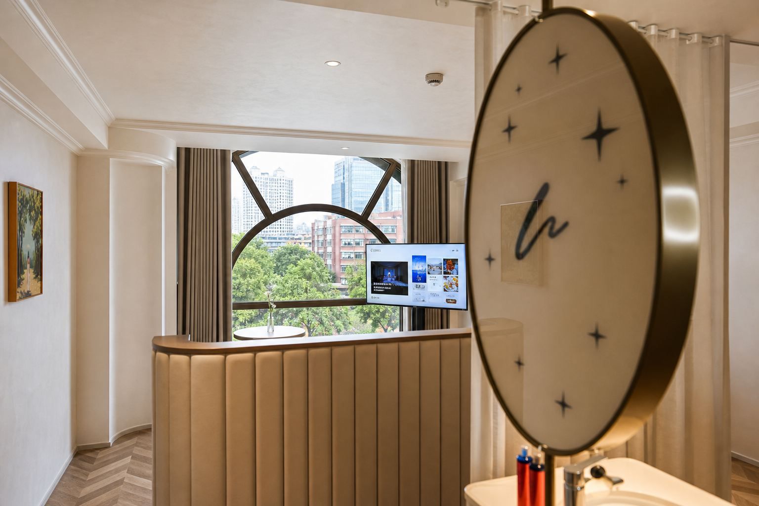Quarto de hotel com uma janela grande, um espelho redondo, uma televisão e vista para edifícios e árvores.