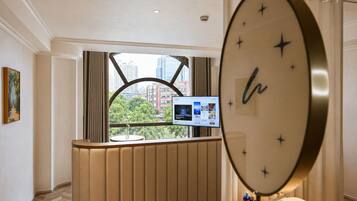 Quarto de hotel com uma janela grande, um espelho redondo, uma televisão e vista para edifícios e árvores.