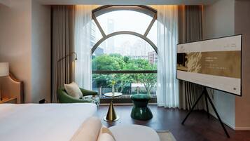 Quarto de hotel com uma cama grande, uma poltrona verde, uma mesinha de centro com um vaso e vista para a cidade através de uma janela ampla.