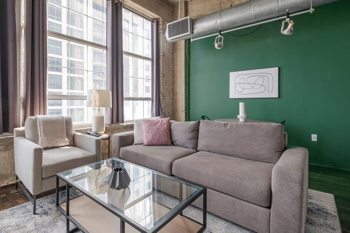 Sala de estar moderna com mesa de centro de vidro, sofá e poltrona. Há uma janela grande com cortinas, uma parede verde com um desenho linear simples e um aparelho de ar condicionado no teto.