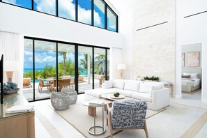 Ampla sala de estar com uma grande janela com vista para uma piscina e vegetação, um sofá branco, uma poltrona estampada e uma mesa de centro com livros.