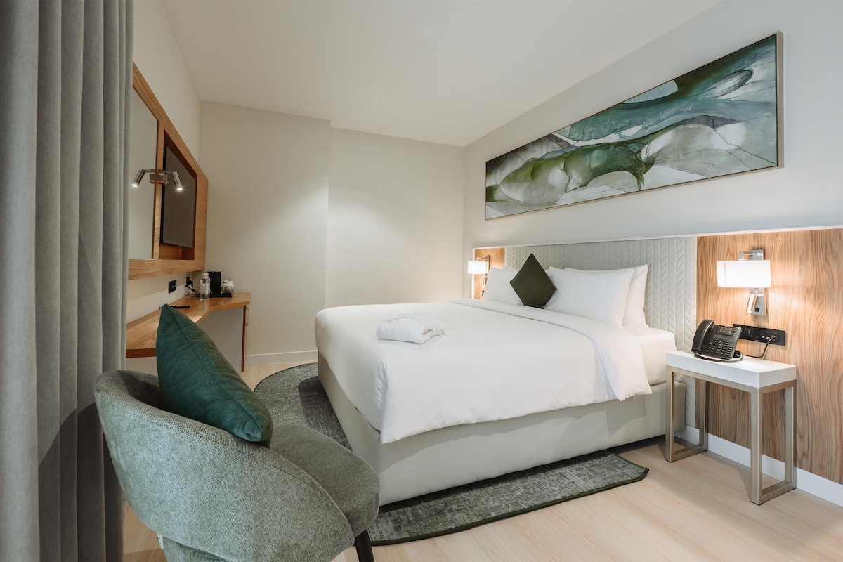 Quarto de hotel com cama, mesa de cabeceira, abajur e um quadro na parede.