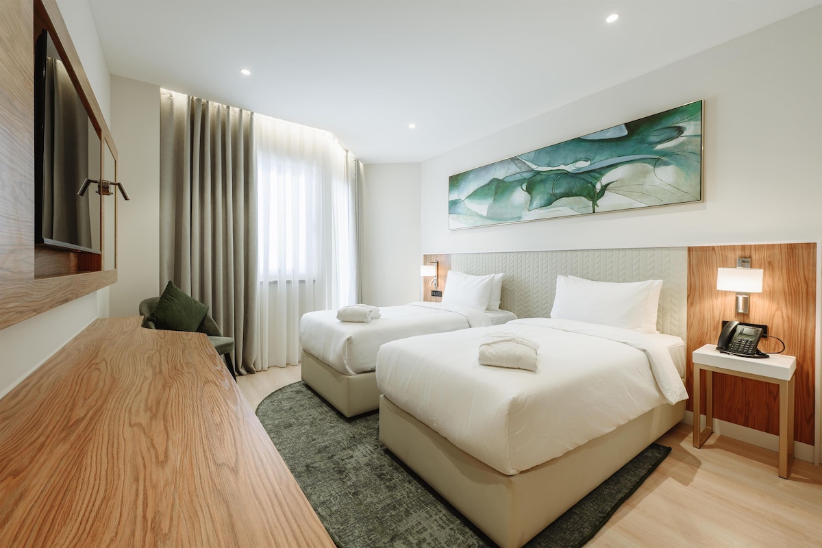 Quarto de hotel com duas camas, uma escrivaninha, uma cadeira, um quadro e um telefone em uma mesinha de cabeceira.