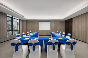 Uma sala de conferências com uma mesa comprida, cadeiras brancas adornadas com fitas azuis e uma tela de projetor na parede.