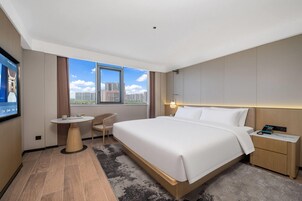 Quarto de hotel com uma cama grande, escrivaninha, cadeira, televisão e vista para a paisagem urbana.