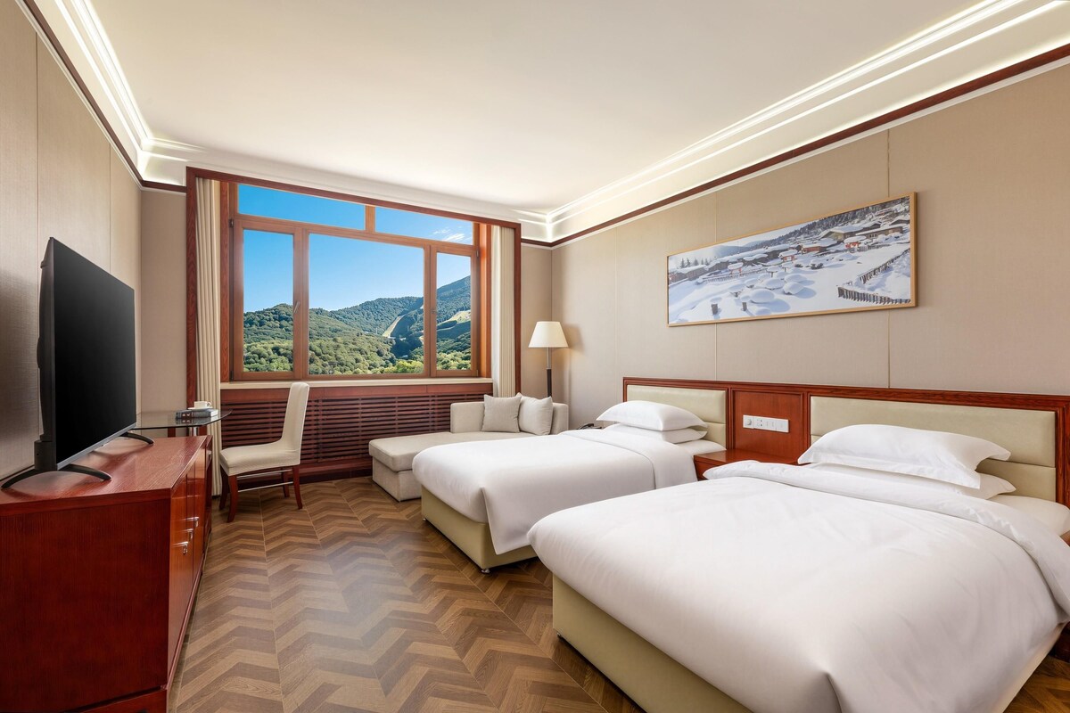 Quarto de hotel com duas camas, escrivaninha, televisão e janela com vista para uma paisagem montanhosa.
