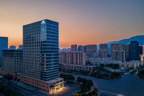 Um hotel moderno e imponente com fachada de vidro, rodeado por paisagens urbanas ao entardecer.