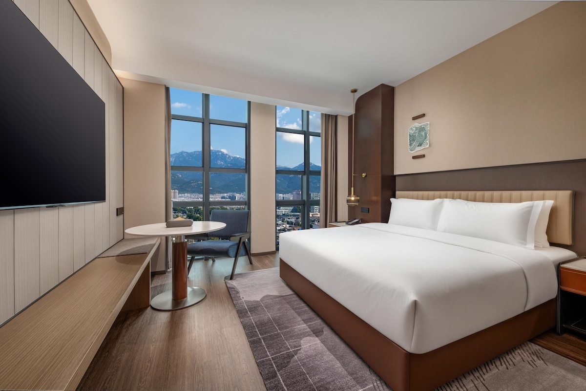 Quarto de hotel com uma cama grande, uma escrivaninha com cadeira, uma TV e vista para a paisagem urbana através de amplas janelas.