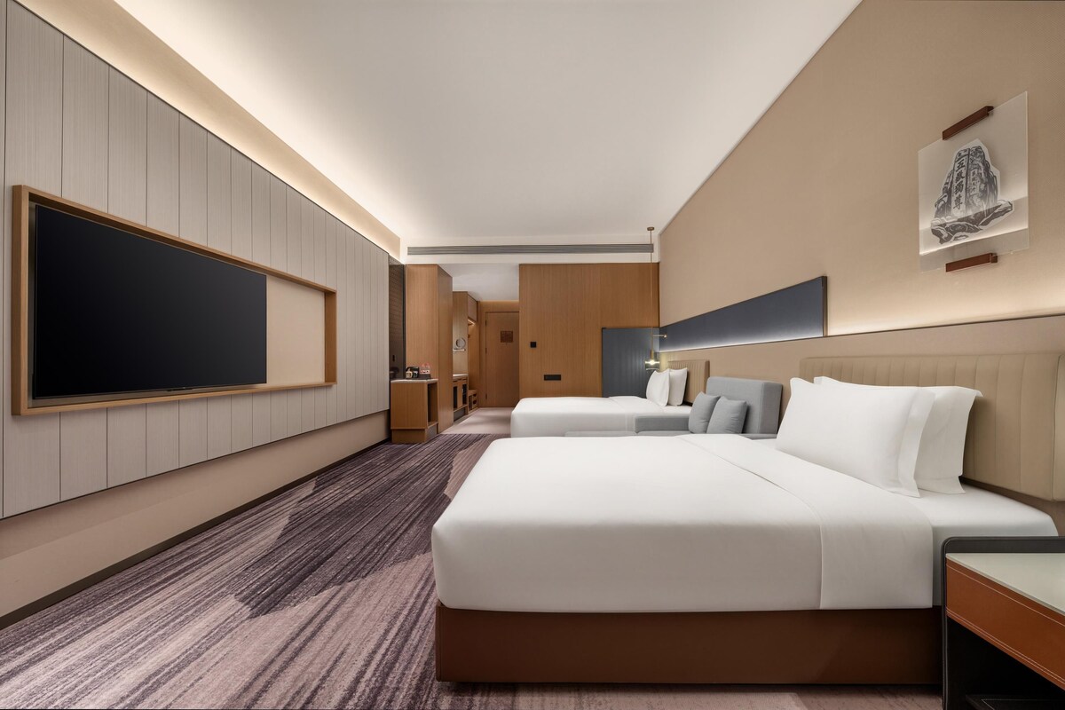 Quarto de hotel com televisão de tela grande, duas camas, escrivaninha e uma obra de arte na parede.