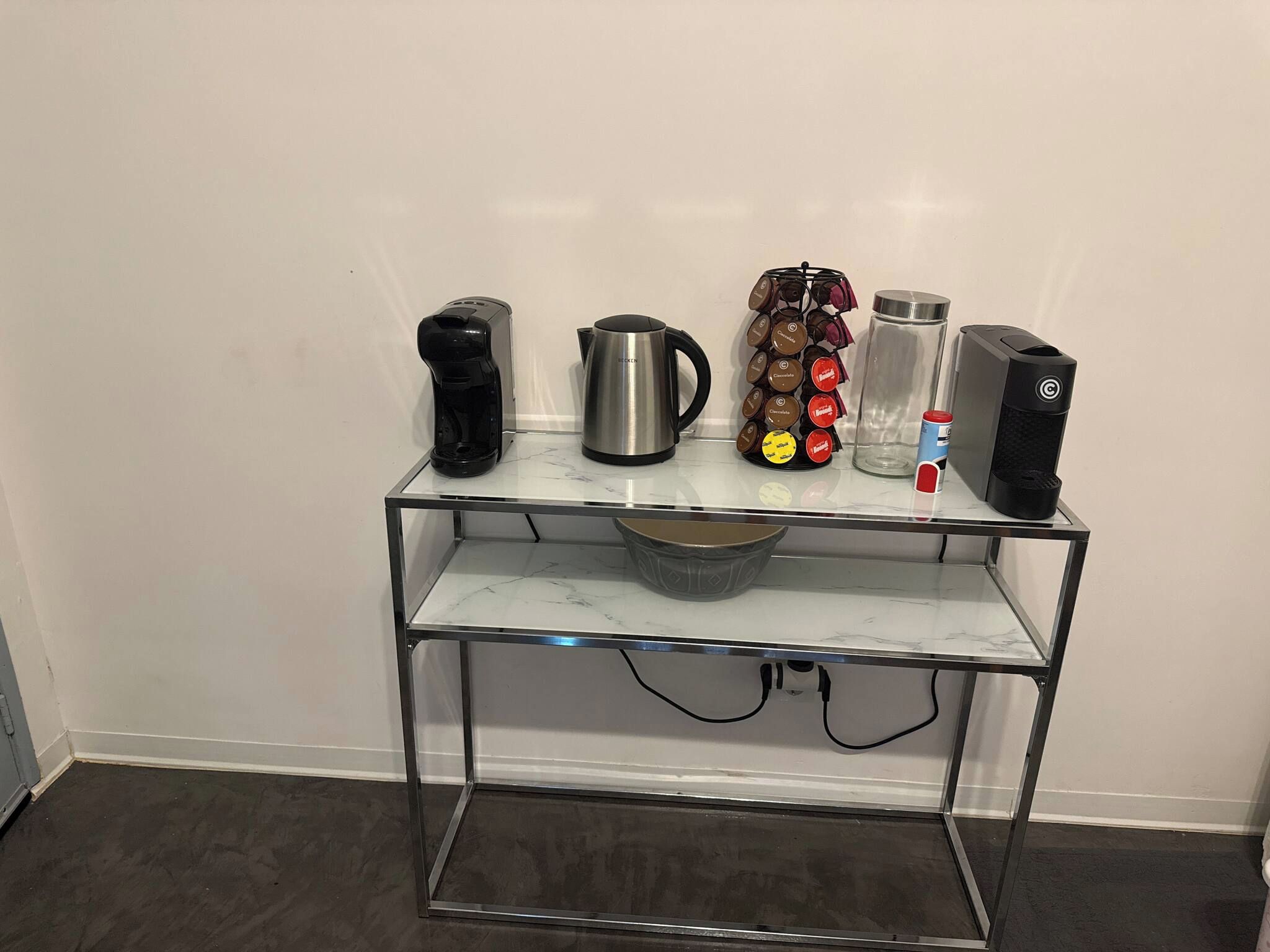 Uma bancada de cozinha com cafeteira, chaleira, caneca, suporte para cápsulas de café, copo e um alto-falante.