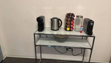 Uma bancada de cozinha com cafeteira, chaleira, caneca, suporte para cápsulas de café, copo e um alto-falante.