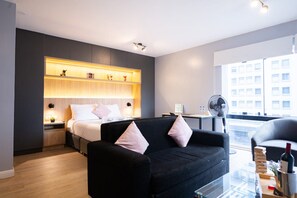 Quarto de hotel moderno com uma cama, um sofá preto, uma mesa de cabeceira com abajur, um ventilador e uma janela com vista para prédios.
