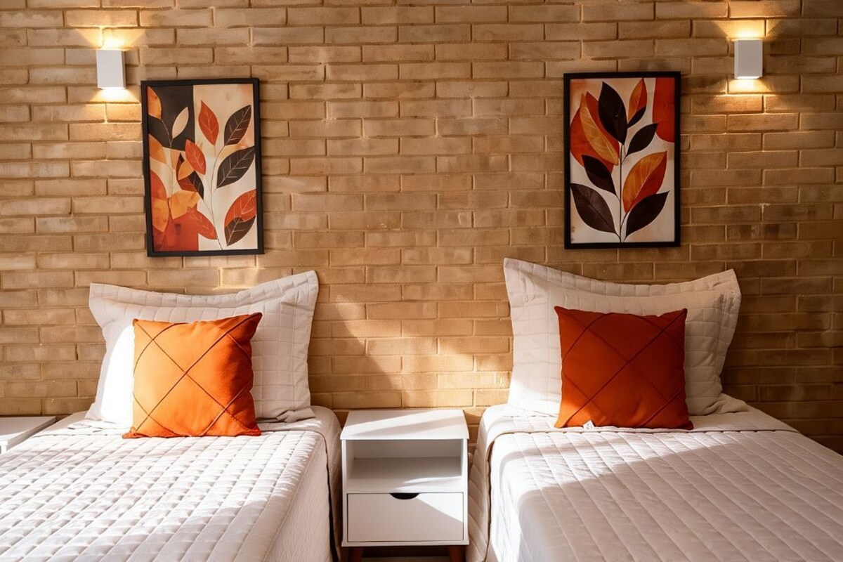 Quarto de hotel com duas camas, cada uma com roupa de cama branca e almofadas laranja, ladeadas por obras de arte penduradas na parede.