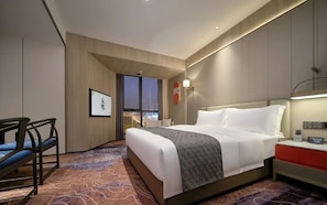 Quarto de hotel com cama, mesa de cabeceira, cadeira e vista para a paisagem urbana pela janela.
