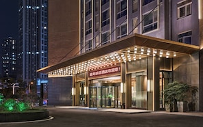 Entrada de hotel com design moderno, fachada em vidro e placa de boas-vindas.