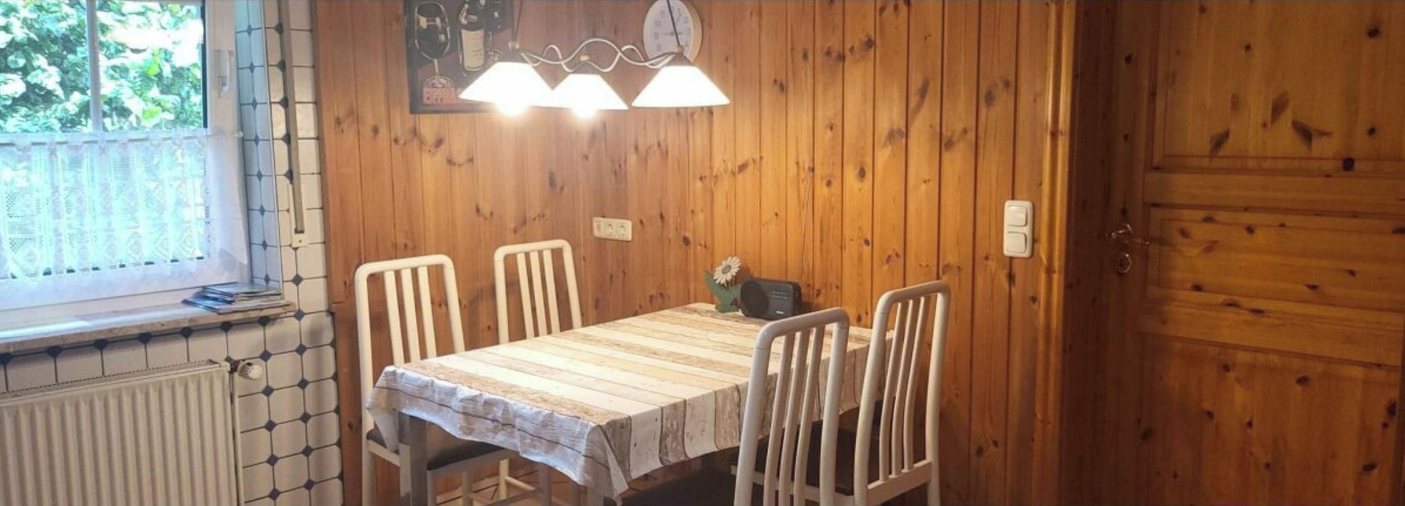 Um quarto com painéis de madeira, uma mesa, cadeiras, uma janela com vista e um relógio na parede.