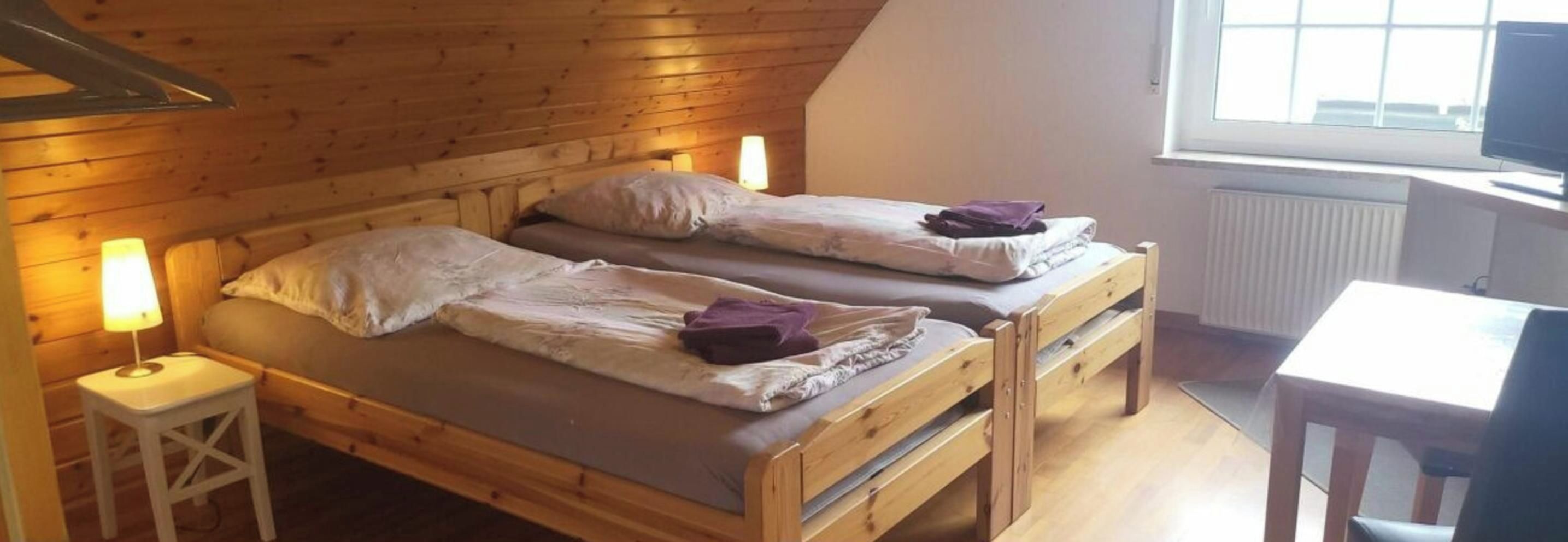 Quarto com duas camas de madeira, cada uma com travesseiro e toalha dobrada. Há uma pequena mesa e uma cadeira à direita, e uma janela que proporciona luz natural.