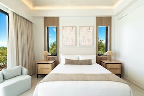 Quarto de hotel moderno com uma cama grande, duas mesas de cabeceira, um criado-mudo, um sofá e janelas amplas com cortinas.