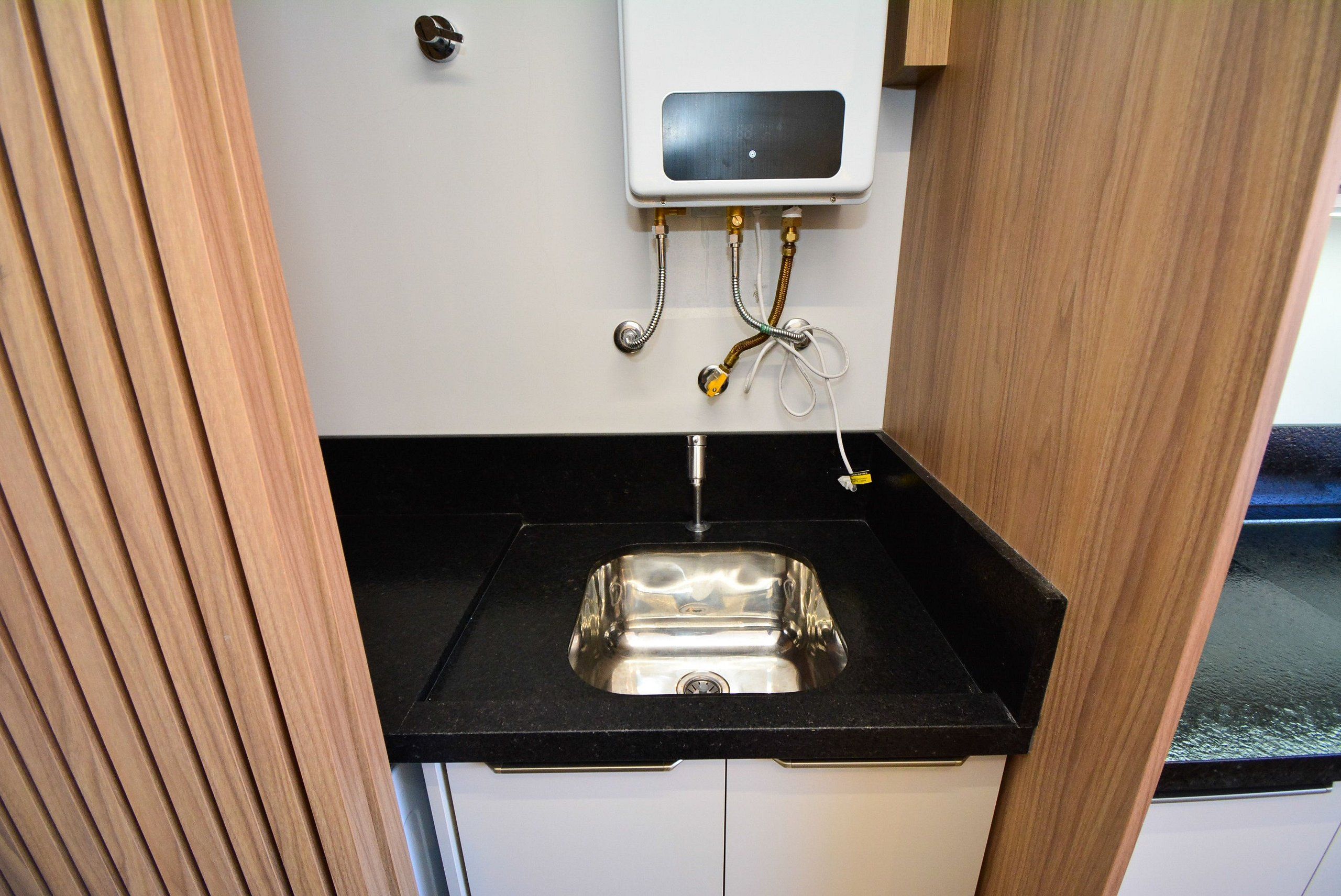 Cozinha compacta com pia de aço inoxidável, armário branco pequeno e aquecedor de água instalado na parede.