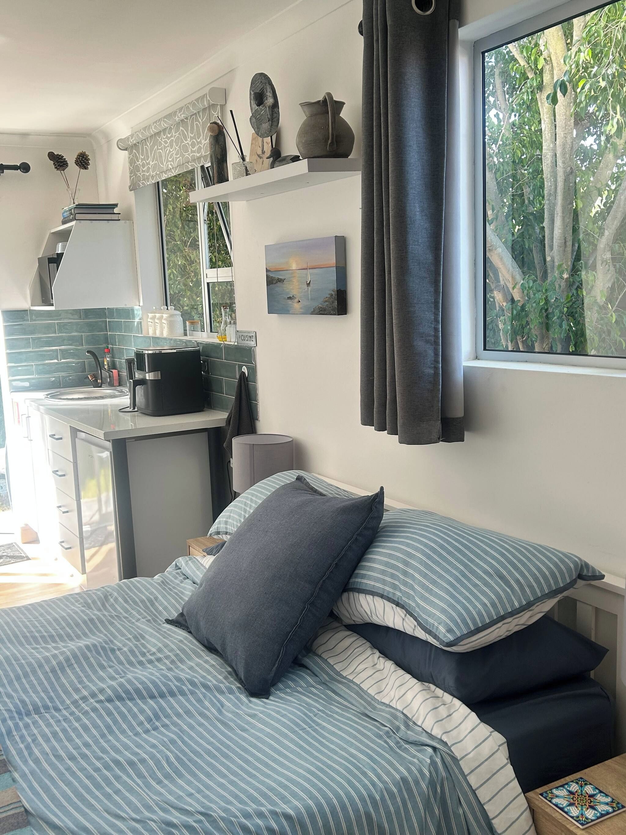 Quarto com cama, almofadas, mesa de cabeceira, janela com cortinas, área de cozinha com fogão e um quadro na parede.