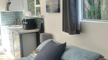 Quarto com cama, almofadas, mesa de cabeceira, janela com cortinas, área de cozinha com fogão e um quadro na parede.