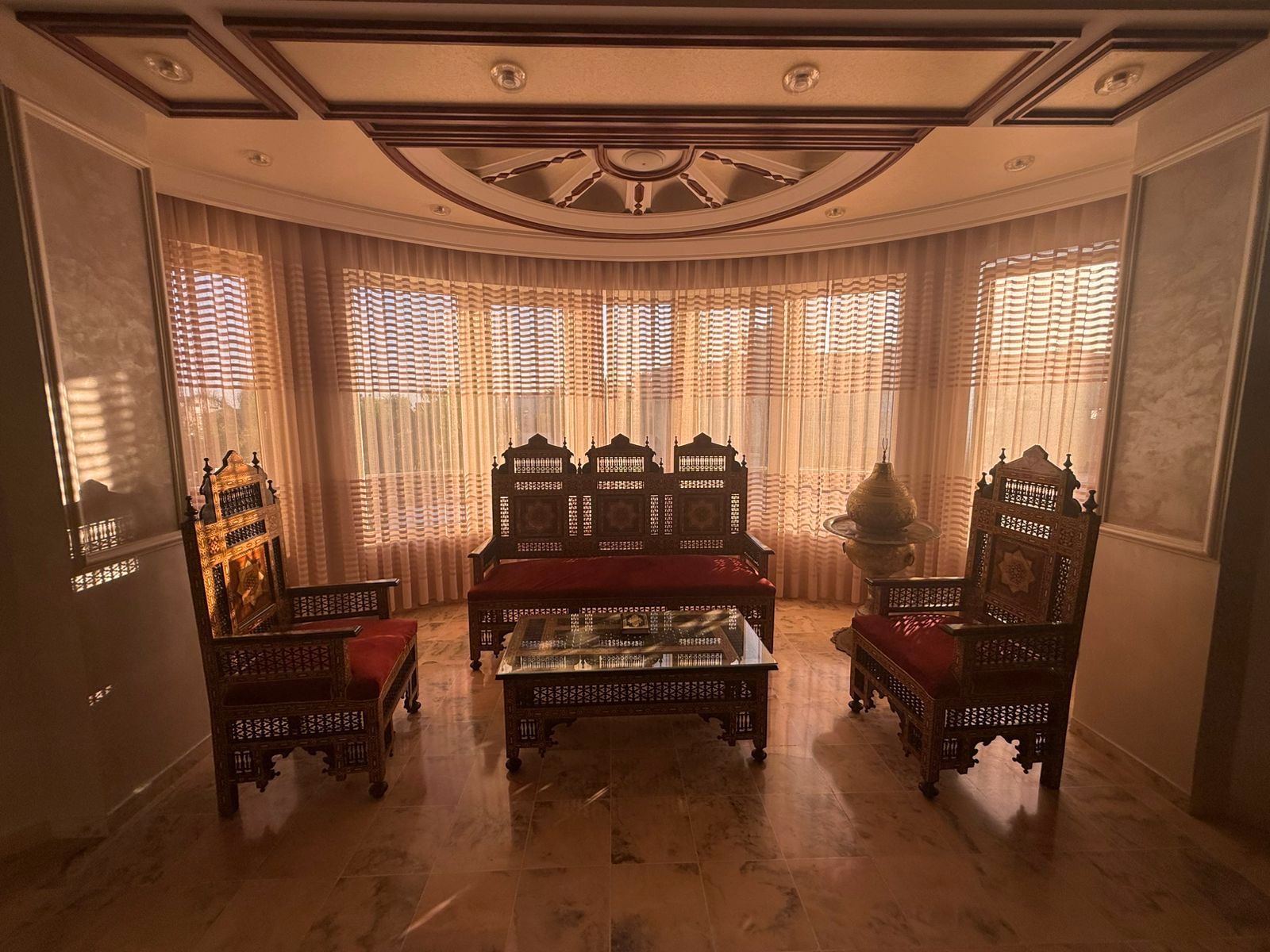 Quarto com mesa central, duas cadeiras ornamentadas e abajur decorativo.