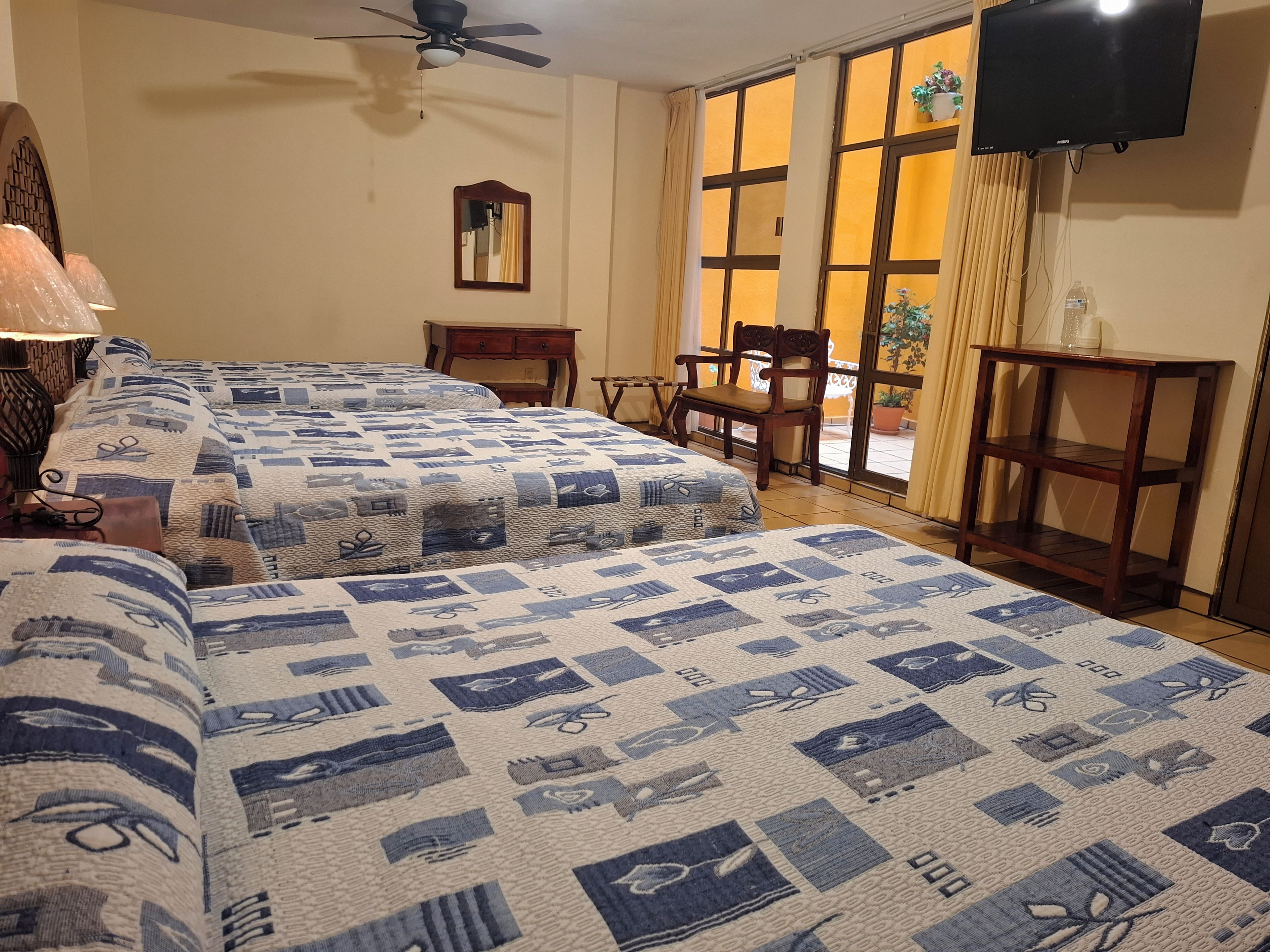 Quarto de hotel com duas camas, ventilador de teto, televisão e vista para o exterior por amplas janelas.