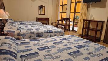 Quarto de hotel com duas camas, ventilador de teto, televisão e vista para o exterior por amplas janelas.