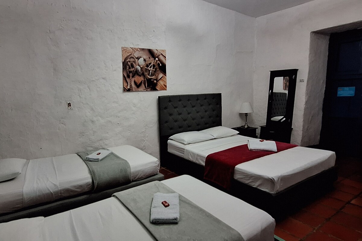 Quarto de hotel com duas camas de solteiro, uma poltrona estofada em verde, um quadro na parede e uma porta que dá para outro cômodo.