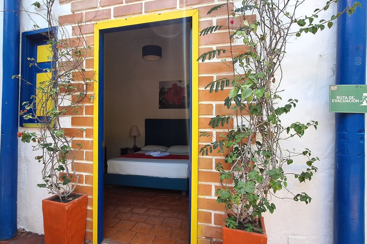 Uma porta dá acesso a um quarto com uma cama, mesa de cabeceira e luminária na parede. Há uma placa verde indicando uma rota de fuga.