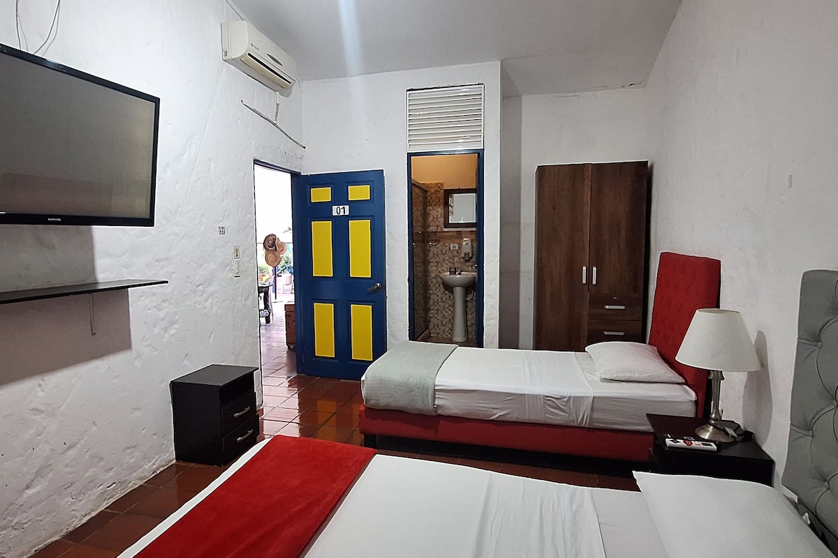 Quarto de hotel com cama, mesa de cabeceira, abajur, guarda-roupa e televisão na parede.
