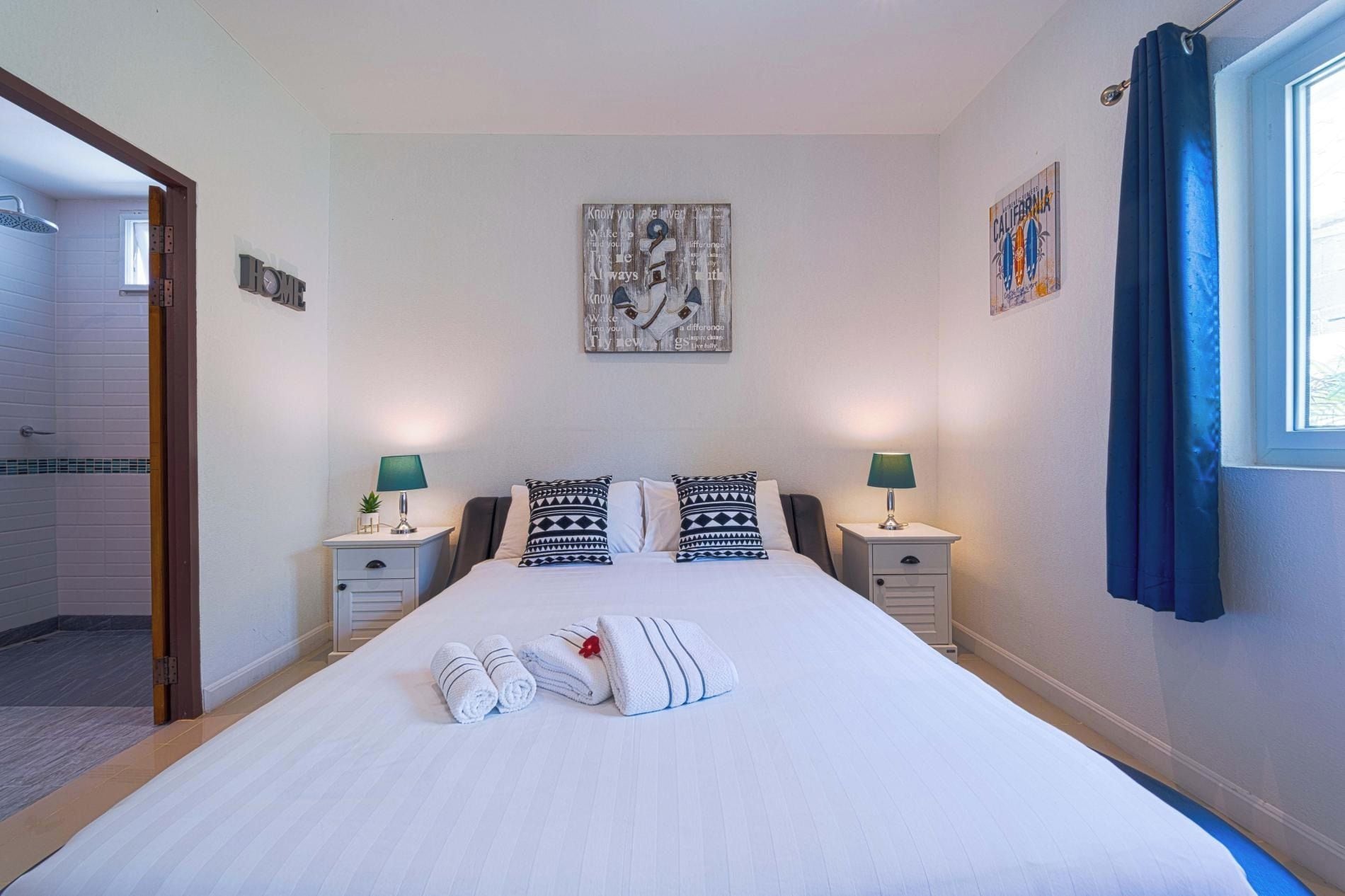 Quarto de hotel com cama, duas mesas de cabeceira, um criado-mudo, uma janela com cortinas e uma porta para um banheiro.