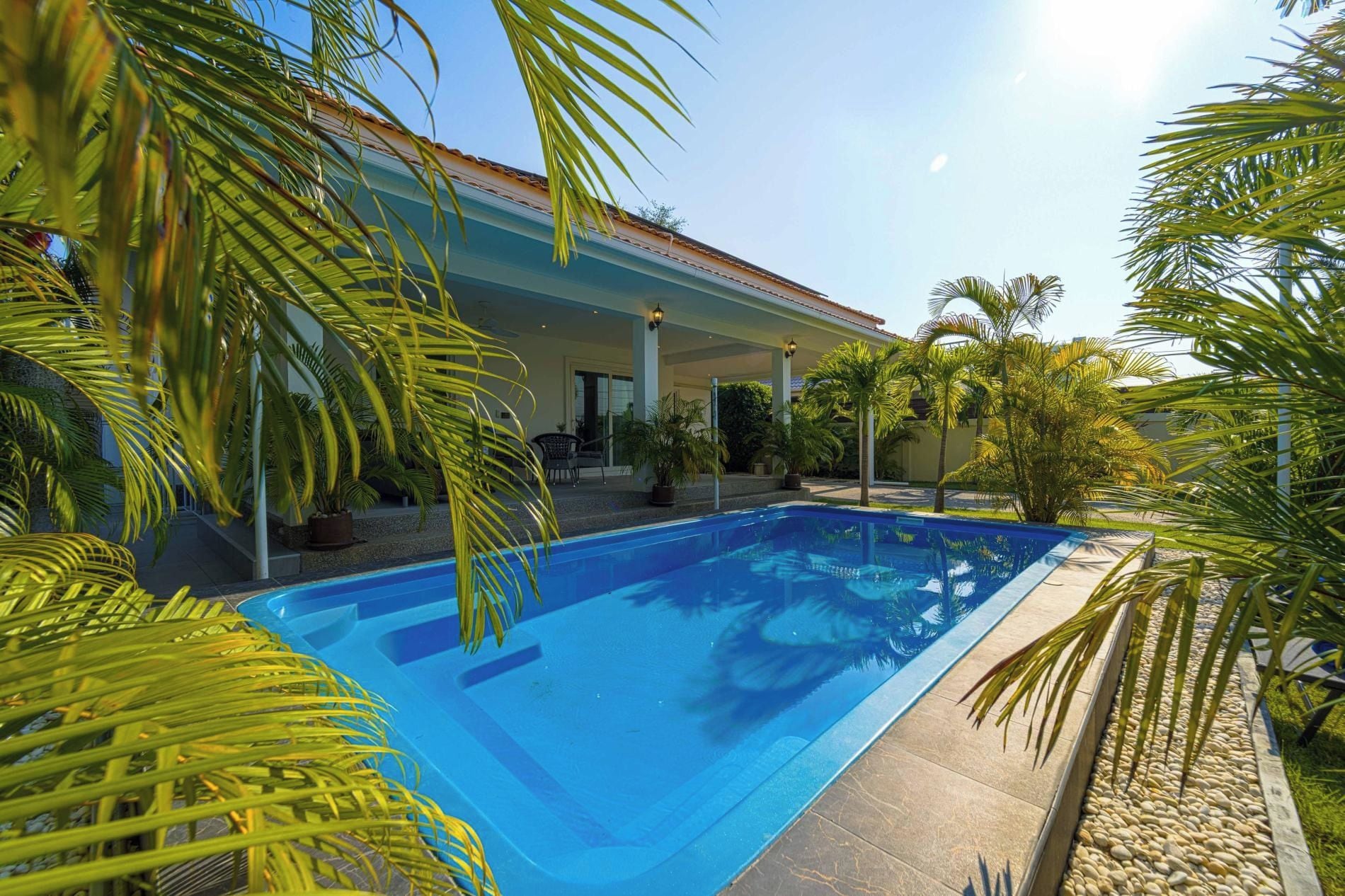 Piscina com deck revestido, rodeada por palmeiras e um espaço coberto com varanda.