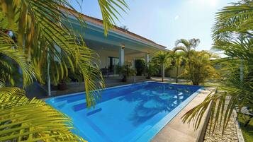 Piscina com deck revestido, rodeada por palmeiras e um espaço coberto com varanda.