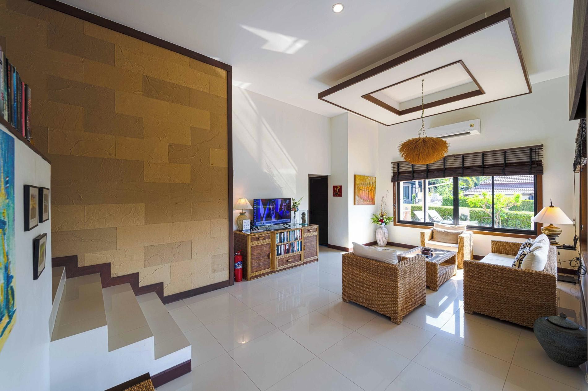 Uma sala de estar ampla, com teto alto, televisão, mobiliário de vime e uma grande janela com vista para o exterior.