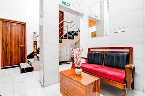 Um saguão de hotel com uma porta de madeira com a placa '101', uma escadaria, um sofá com almofadas vermelhas e uma pequena mesa com um vaso.
