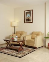 Uma sala de estar com dois poltronas bege, uma mesa de centro, um abajur e um quadro na parede.