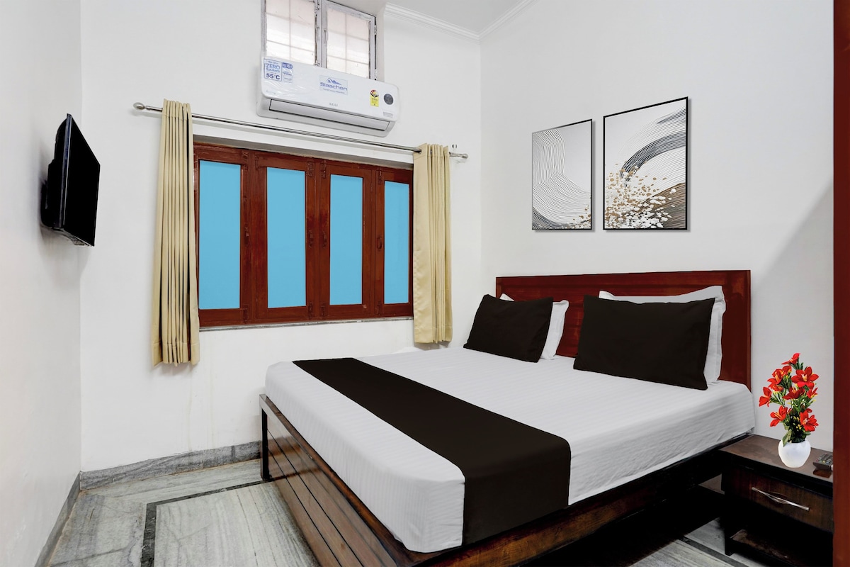 Quarto de hotel com cama, mesa de cabeceira, um vaso com flores, janela com cortinas e duas obras de arte nas paredes.
