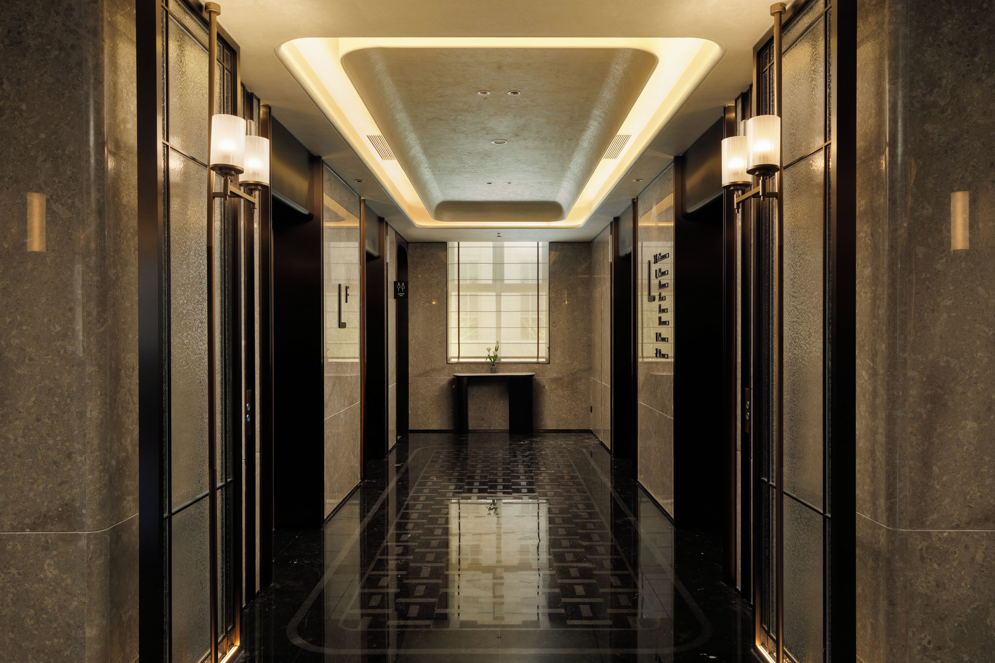 Hall de elevadores moderno com piso refletivo, luminária central e porta de vidro com placa.