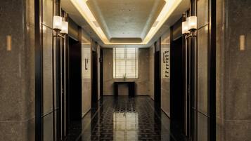 Hall de elevadores moderno com piso refletivo, luminária central e porta de vidro com placa.