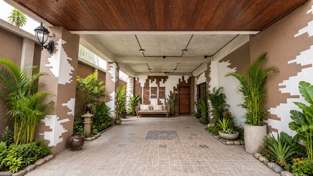 Área externa coberta com piso de azulejos, plantas em vasos, um banco de madeira e uma parede com padrão geométrico.