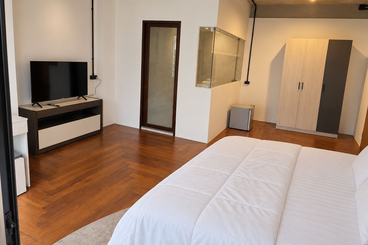 Quarto moderno com uma cama grande, uma televisão em um suporte de madeira, um guarda-roupa e uma porta de vidro que dá para um banheiro.