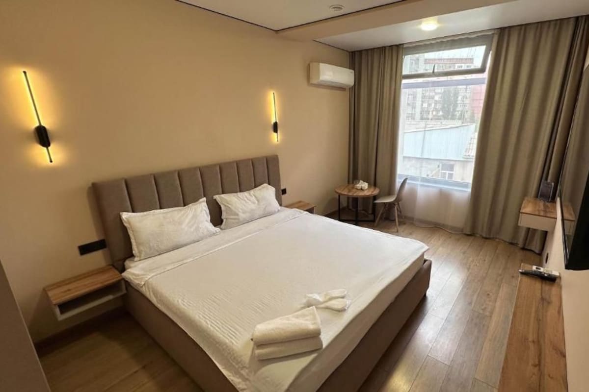 Quarto de hotel com uma cama grande, mesas de cabeceira, uma pequena mesa com cadeiras, uma televisão presa à parede e uma janela com cortinas.