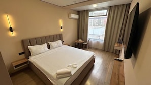Quarto de hotel com uma cama grande, mesas de cabeceira, uma pequena mesa com cadeiras, uma televisão presa à parede e uma janela com cortinas.
