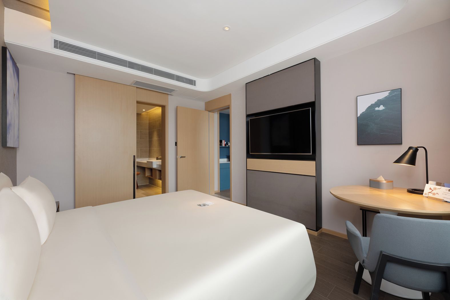 Quarto de hotel moderno com uma cama grande, uma escrivaninha com cadeira, televisão de tela plana e um quadro na parede.
