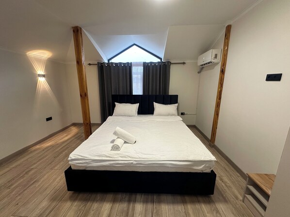 Cama bem arrumada com edredom branco e duas almofadas, piso de madeira, claraboia e ar condicionado fixado na parede.